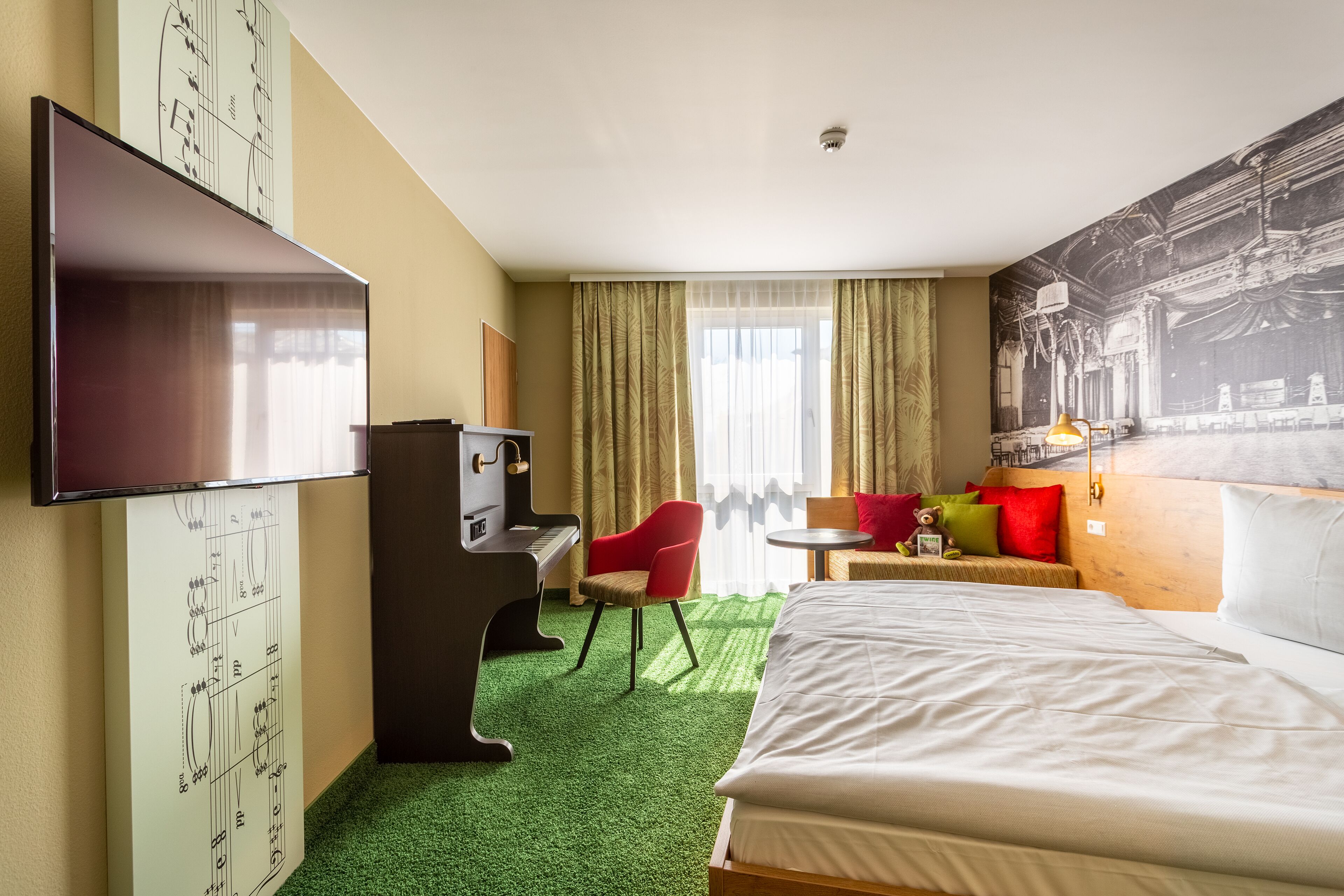 Quarto de hotel com cama, escrivaninha, cadeira, televisão e um mural na parede.