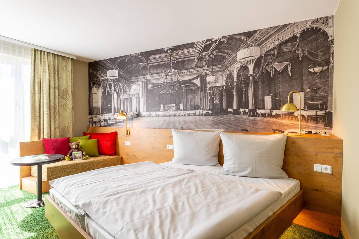 Quarto de hotel com uma cama grande, um sofá pequeno com almofadas coloridas, uma mesa redonda e um mural de um aposento ornamentado na parede.