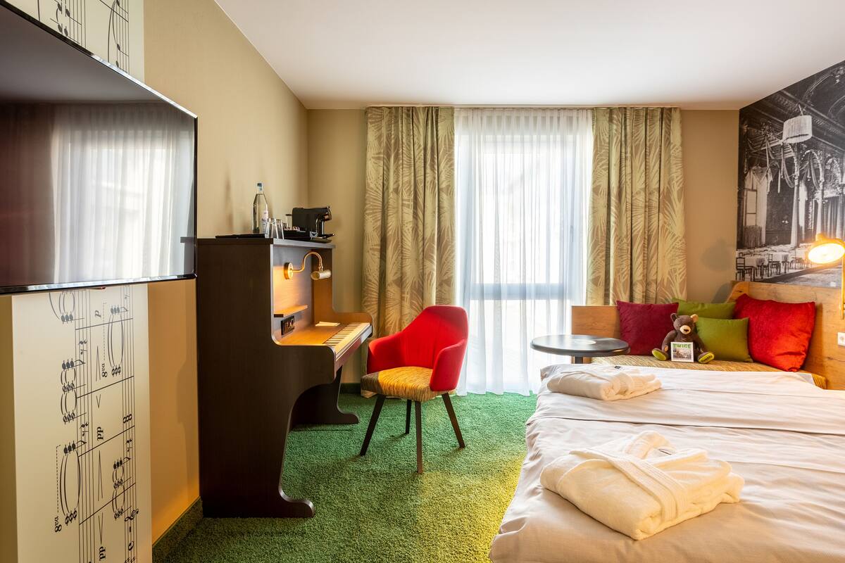 Quarto de hotel com cama, escrivaninha com computador, cadeira e piano.