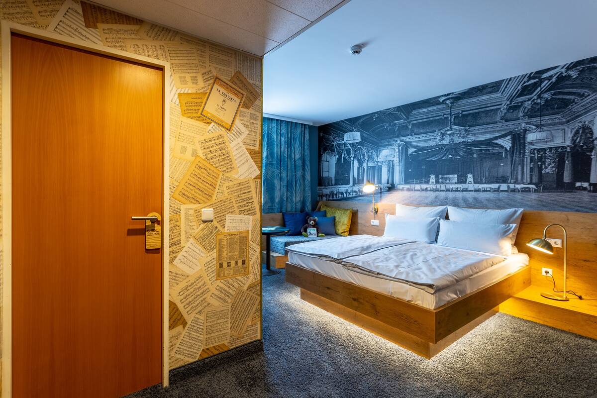 Quarto de hotel com porta de madeira, cama com roupa de cama branca, mesa de cabeceira com abajur e um mural na parede retratando uma cena histórica.