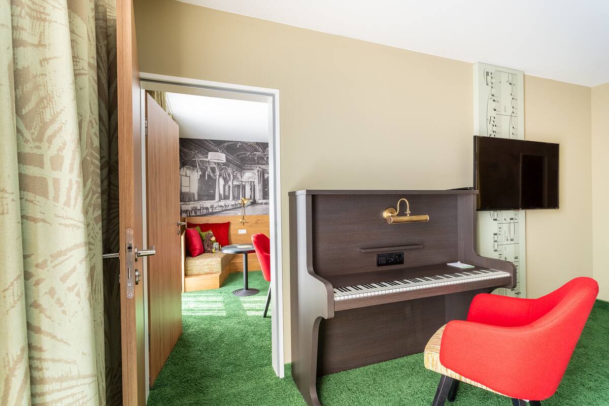 Quarto com piano, uma cadeira vermelha, televisão na parede e porta que dá para outro cômodo.