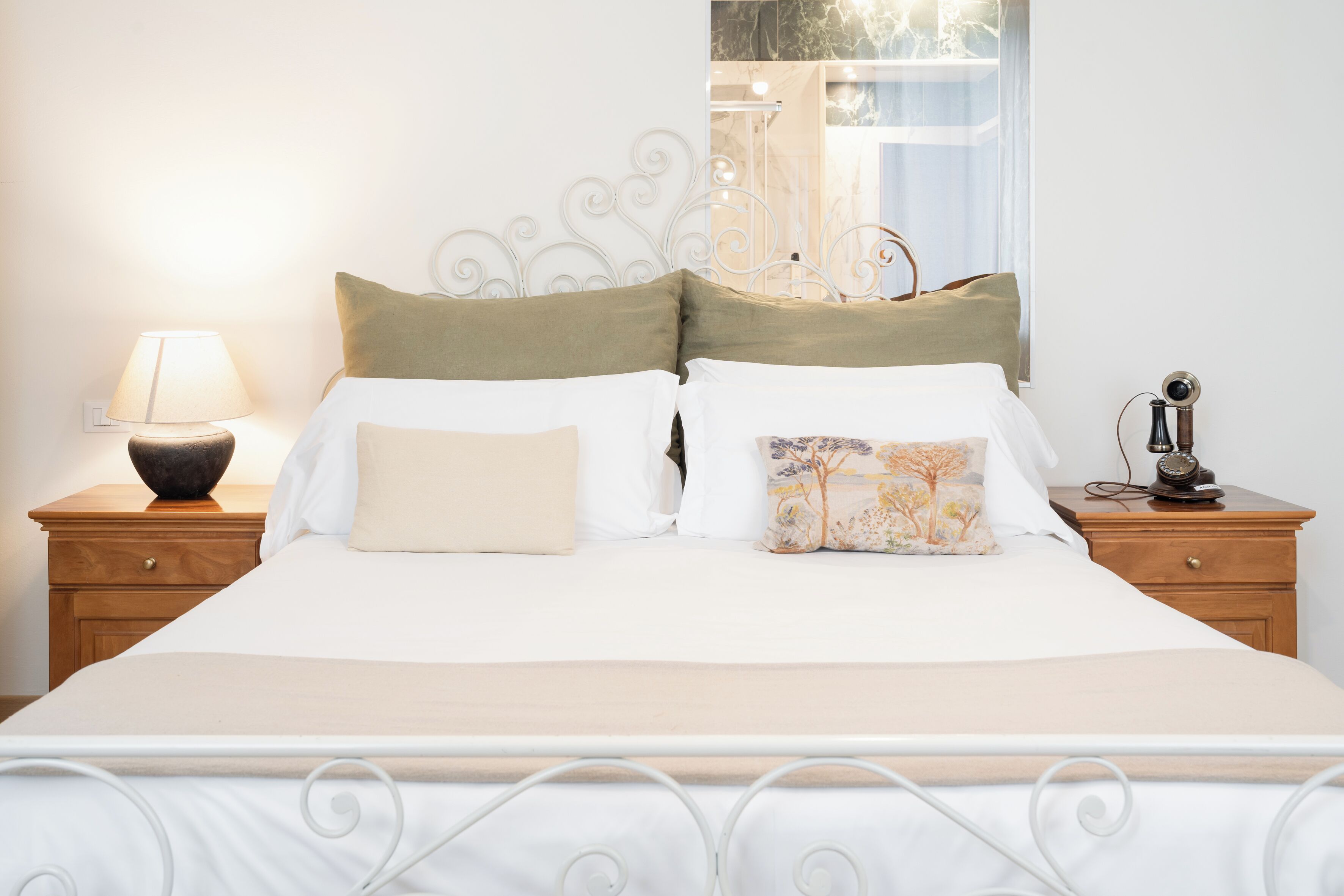 Uma cama bem arrumada com almofadas brancas e verdes, um abajur e um telefone antigo sobre um criado-mudo de madeira.