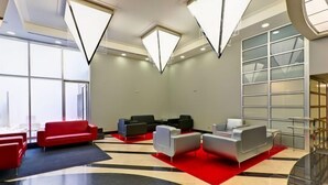 Um lobby moderno com assentos em tons de vermelho e cinza, grandes janelas e luminárias de teto geométricas.