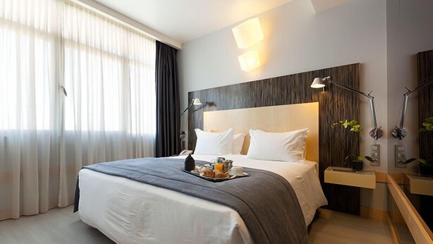 Quarto de hotel moderno com uma cama grande, abajures, mesa de cabeceira e uma janela com cortinas leves.