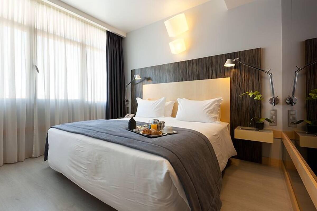 Quarto de hotel moderno com uma cama grande, abajures, mesa de cabeceira e uma janela com cortinas leves.