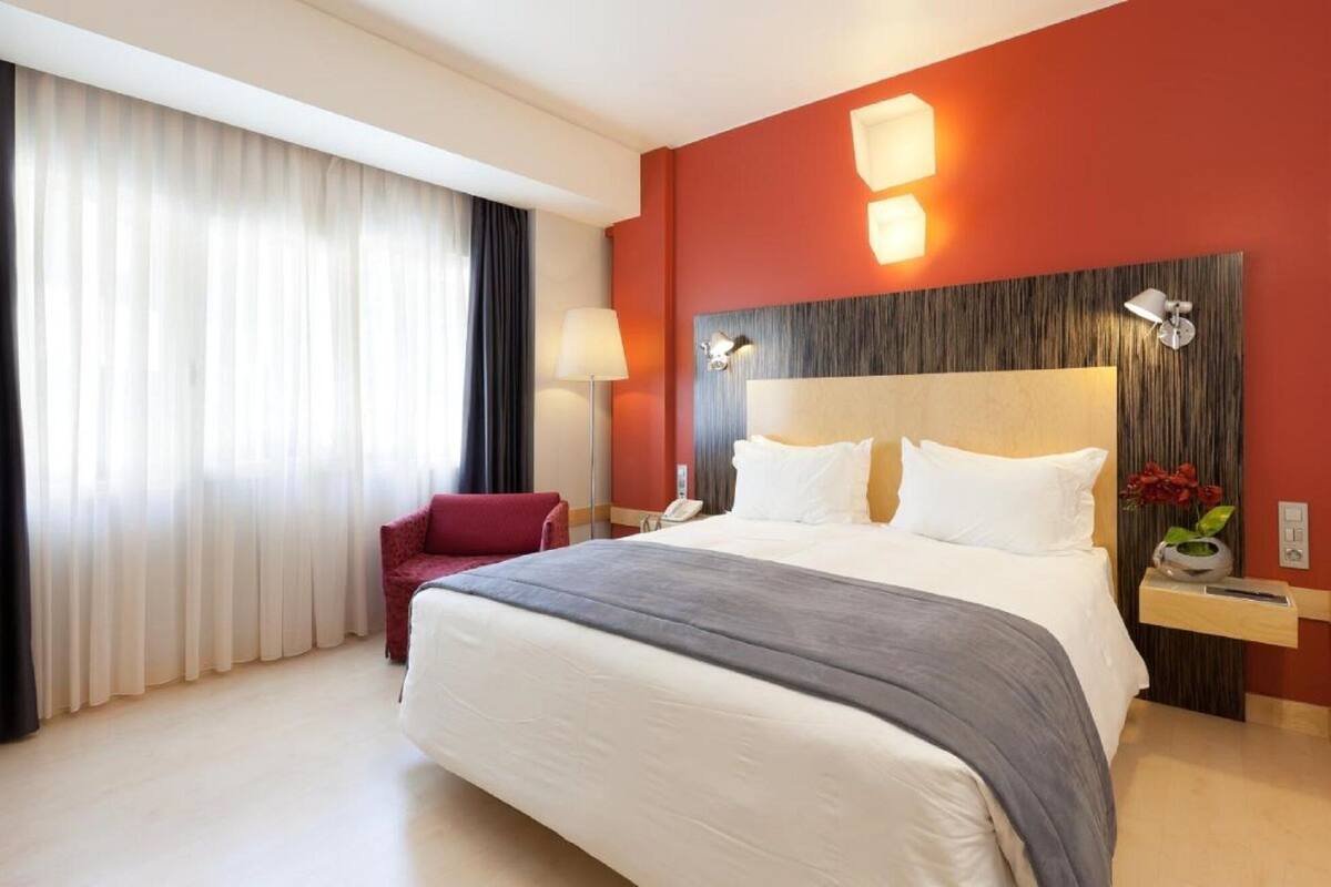 Quarto de hotel com uma cama grande, uma poltrona vermelha, uma mesa de cabeceira com abajur e um vaso com flores.