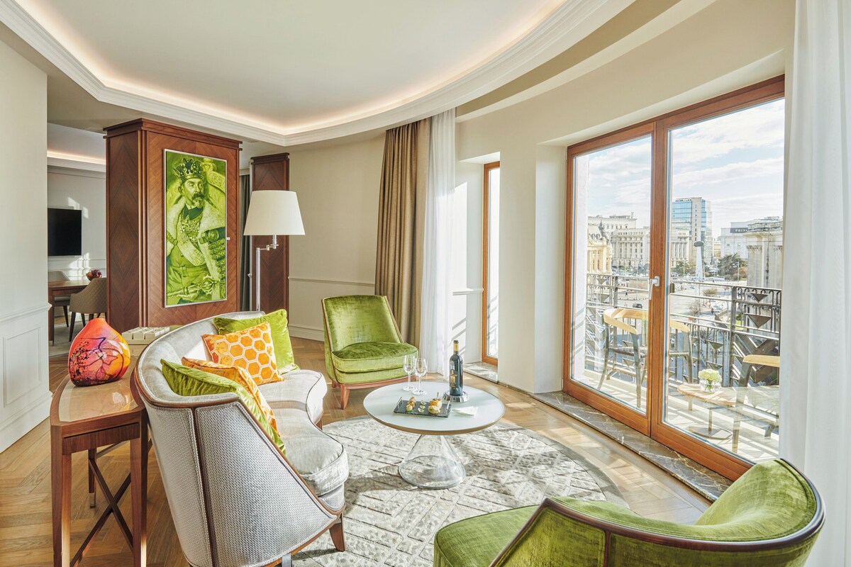 Uma sala de estar ampla com vista para a cidade, uma poltrona verde, um sofá e uma área de jantar com mesa e cadeiras.