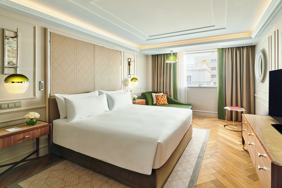 Quarto de hotel com uma cama grande, uma escrivaninha com luminária, uma televisão e vista para a cidade pela janela.