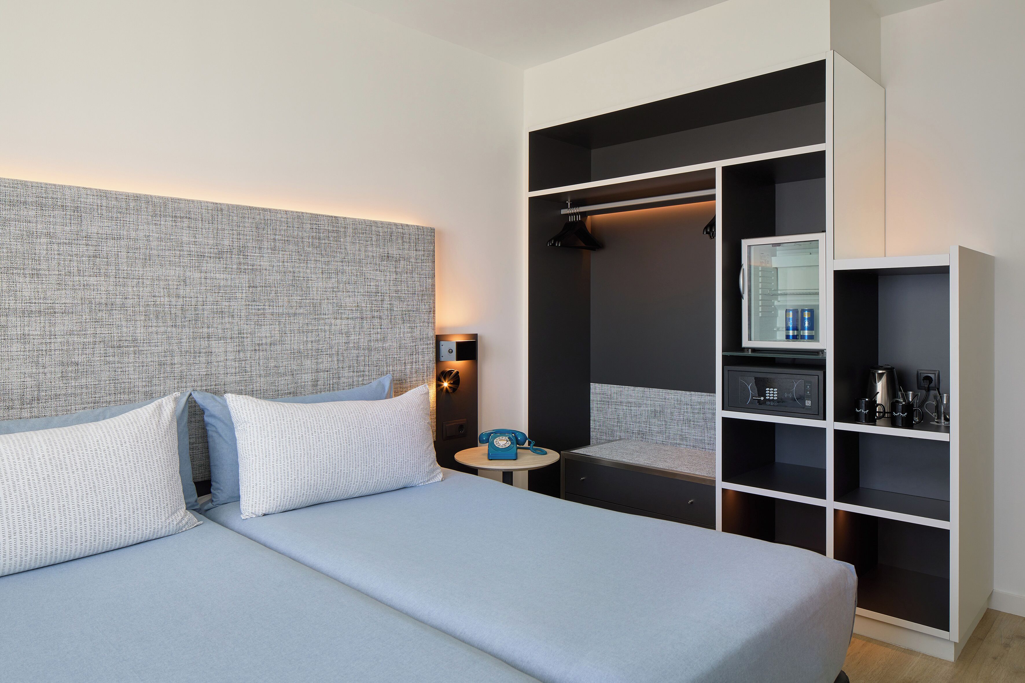 Quarto de hotel moderno com uma cama grande, mesa de cabeceira com abajur, guarda-roupa com espelho e uma pequena mesa com telefone.