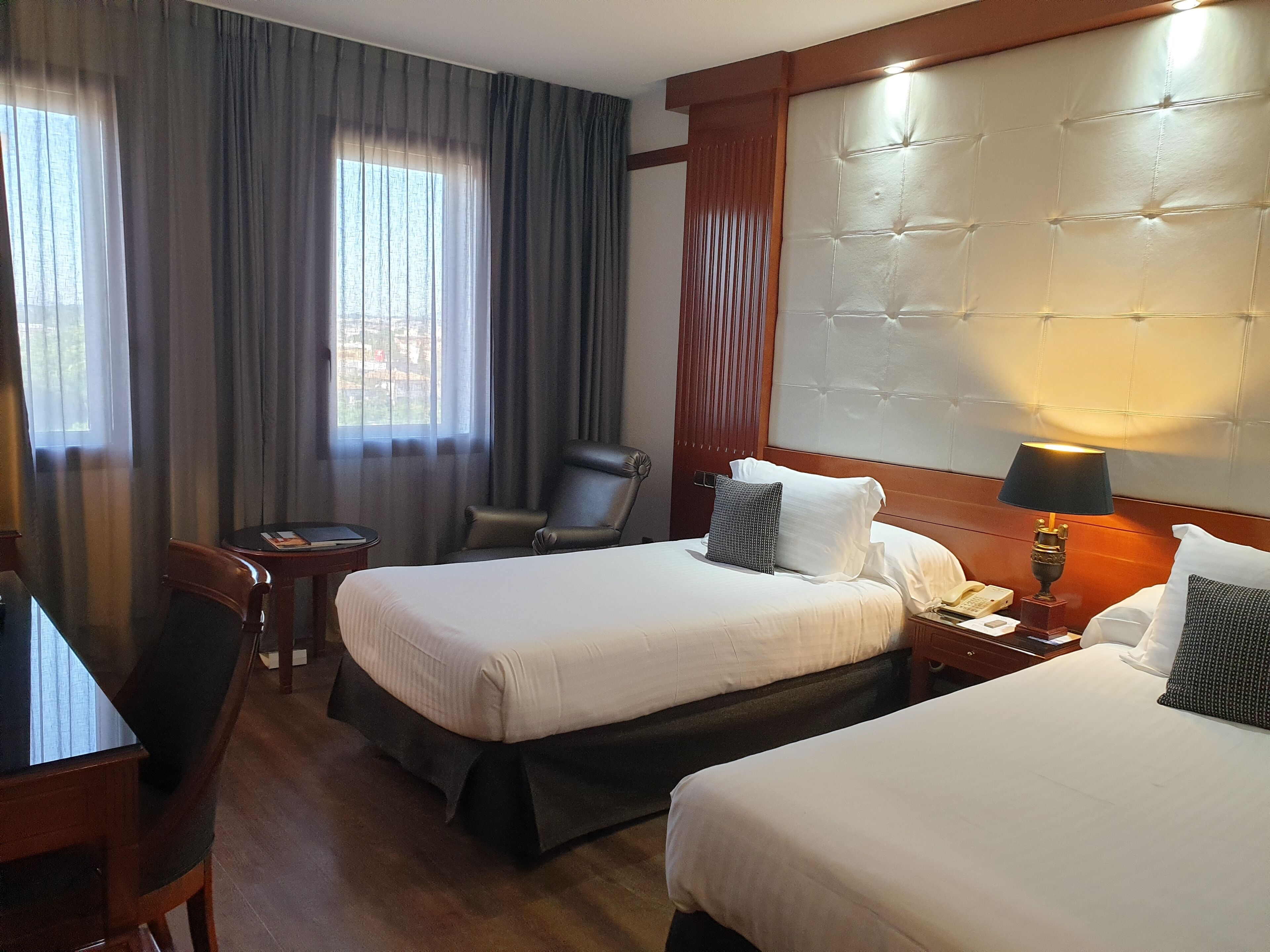 Quarto de hotel com duas camas, uma mesa, uma cadeira e uma janela com cortinas.
