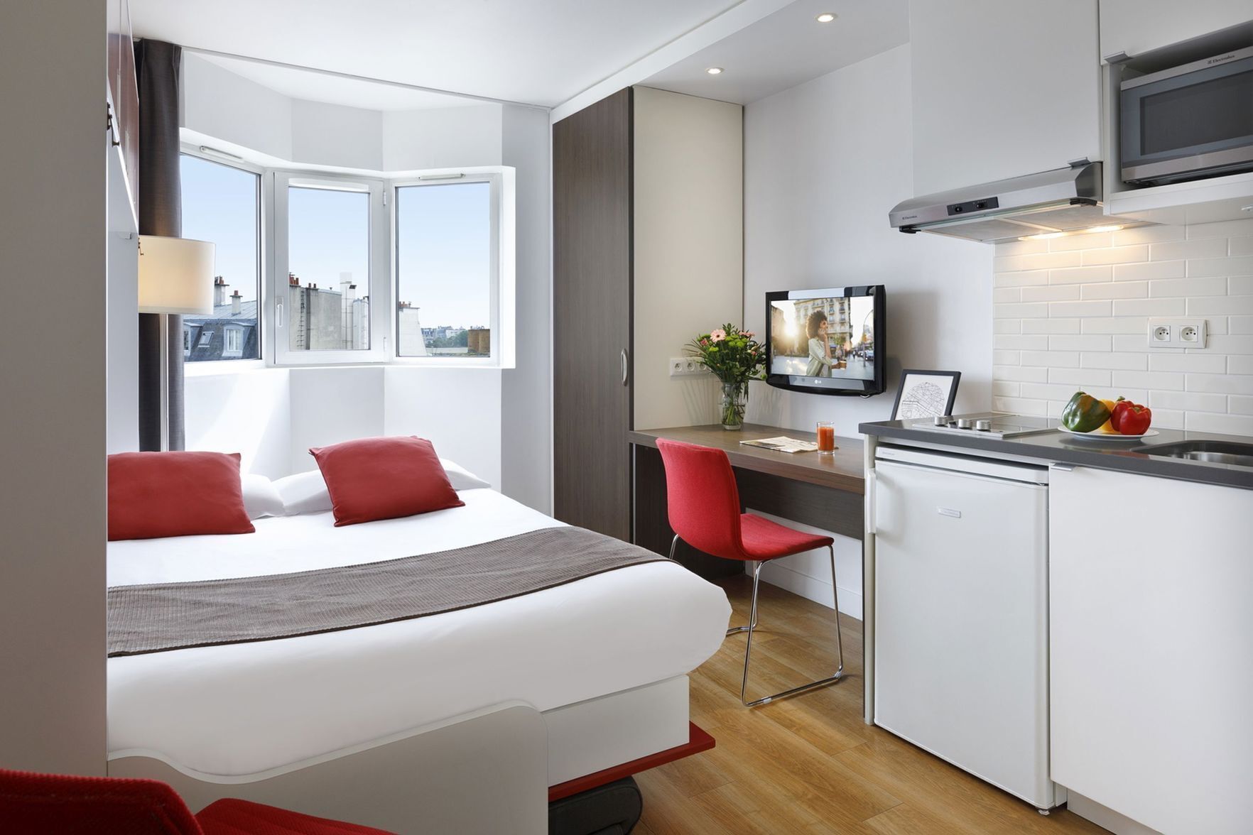 Quarto de hotel compacto com cama, escrivaninha, cadeira, cozinha compacta e uma janela grande.