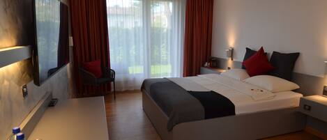 Quarto de hotel moderno com uma cama grande, uma escrivaninha com cadeira, uma TV na parede e uma janela com cortinas.