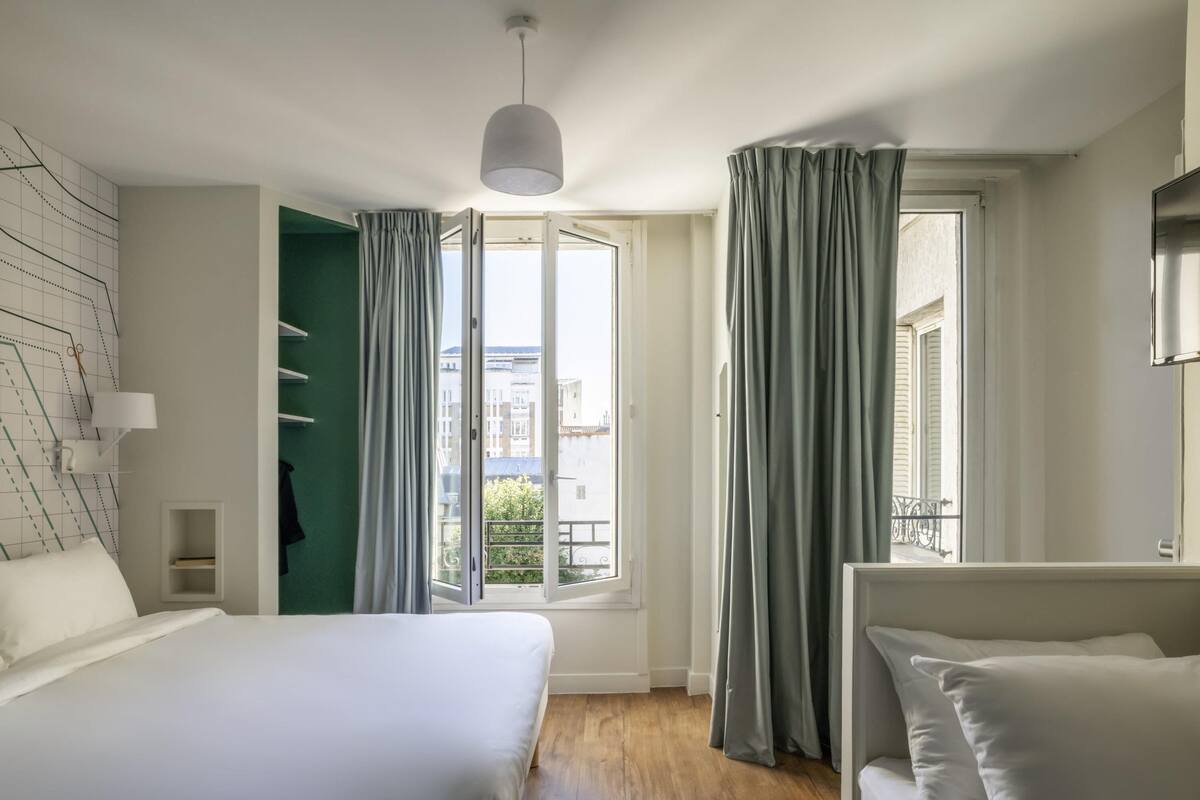 Quarto de hotel moderno com uma cama grande, um sofá, uma janela com cortinas, uma luminária pendente e vista para a cidade.