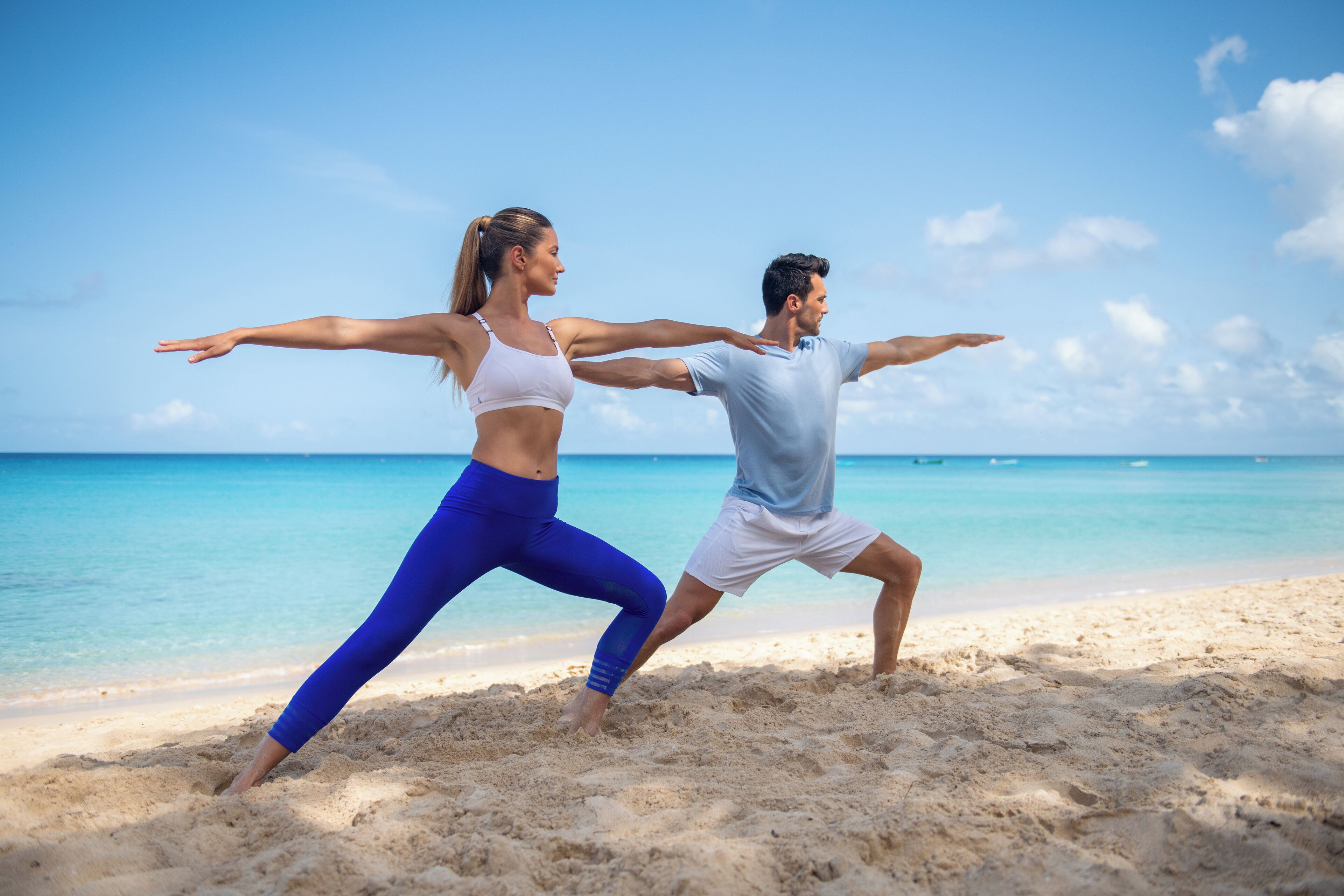 Duas pessoas fazem uma postura de yoga numa praia, com um céu claro e mar calmo ao fundo.