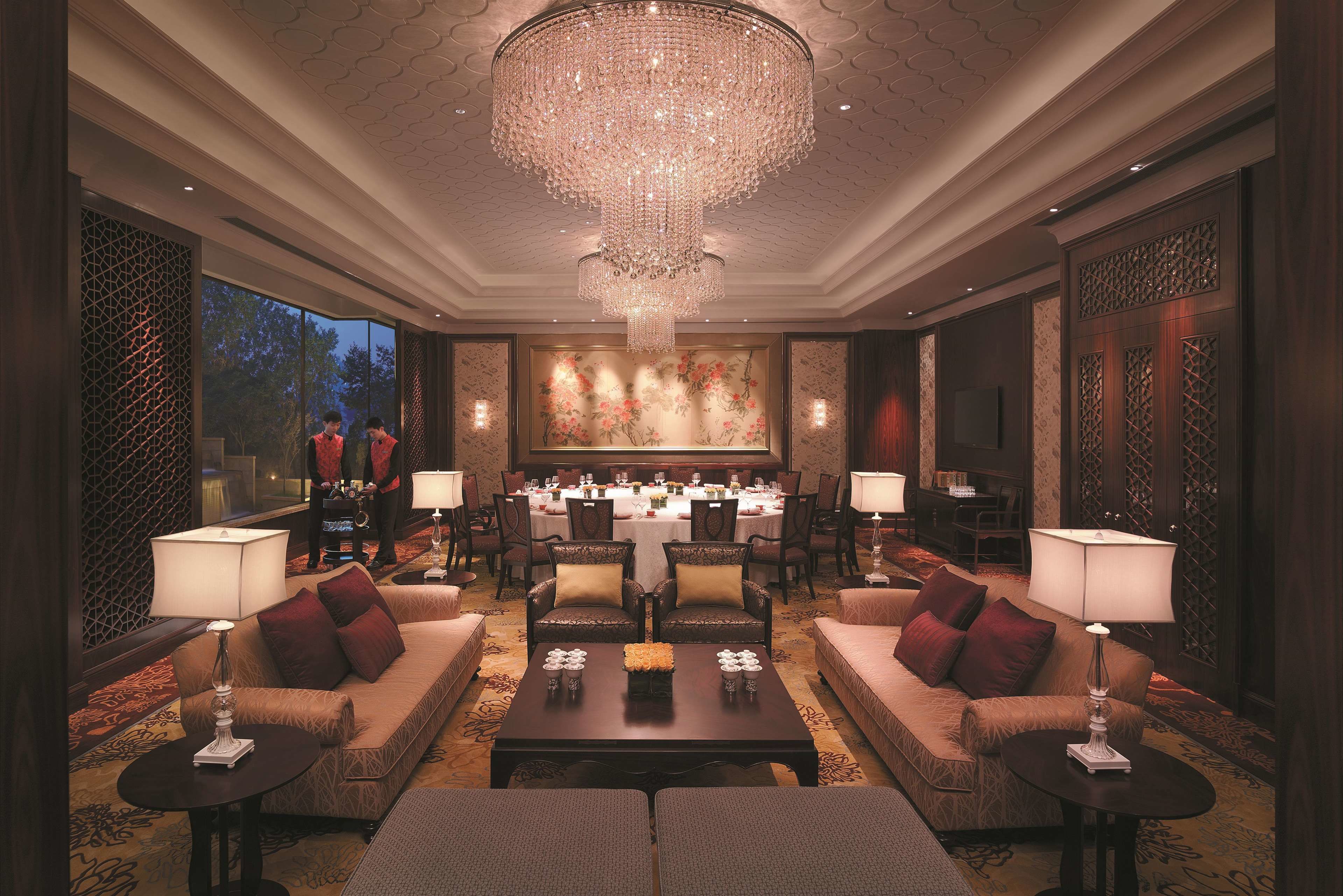 Um hall de hotel luxuoso, com um grande candelabro, assentos confortáveis, área de jantar e uma pintura grande na parede.
