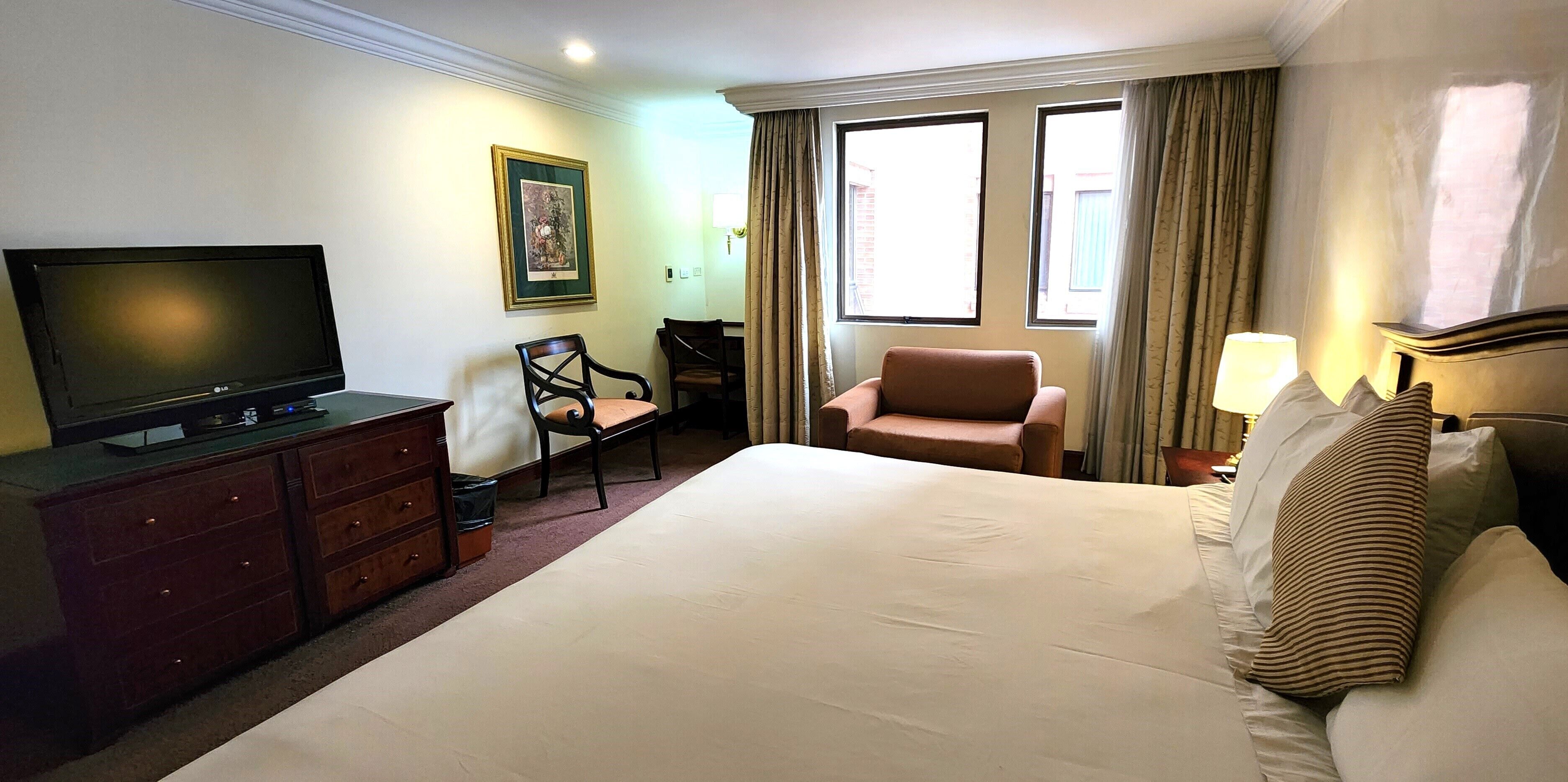 Quarto de hotel com cama, escrivaninha com cadeira, televisão e janela com cortinas.