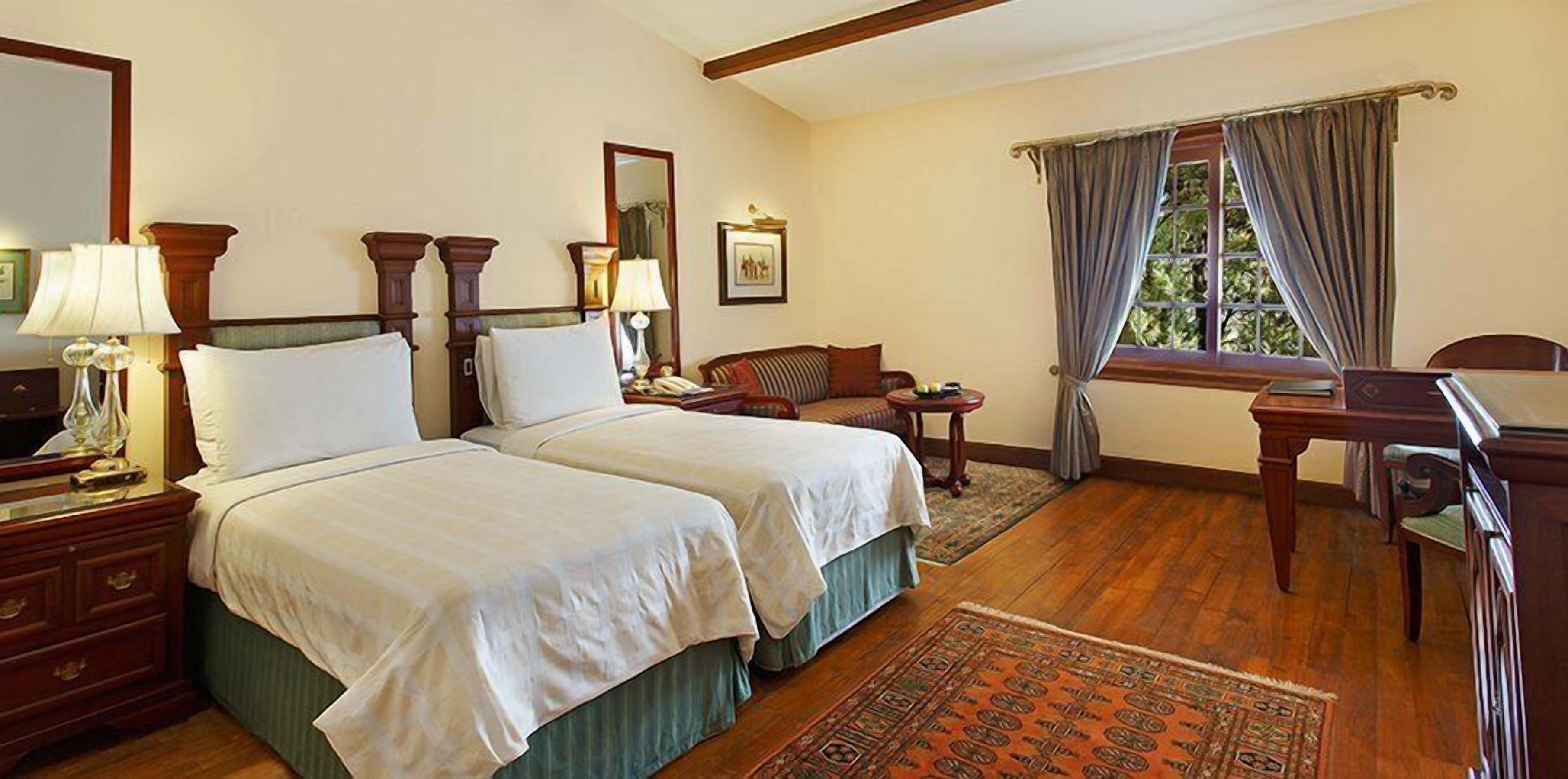 Quarto de hotel com duas camas, uma escrivaninha, uma cadeira, uma mesinha, um abajur, um espelho e uma janela com cortinas.