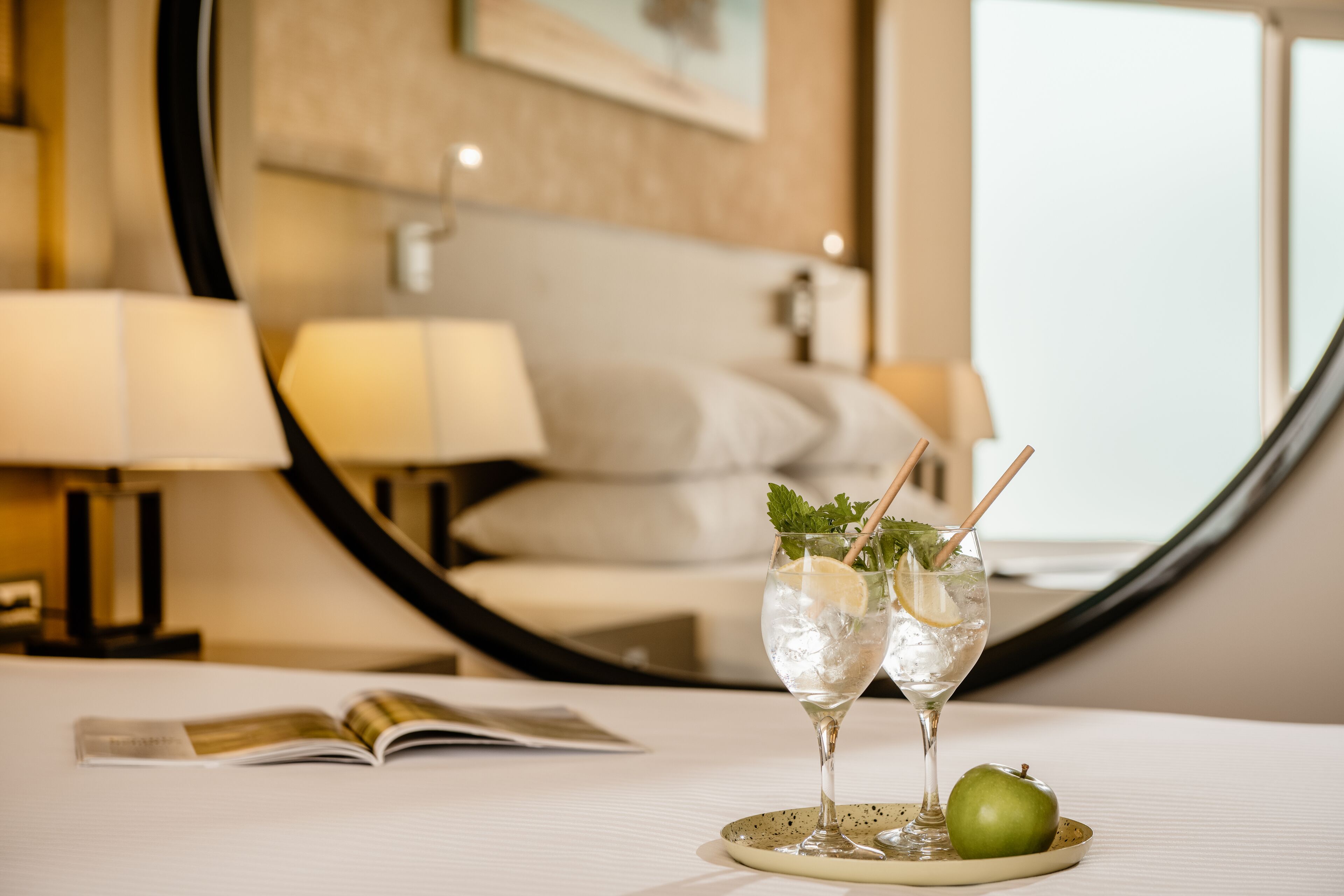 Quarto de hotel bem arrumado, com uma cama, mesa de cabeceira com abajur, e um copo de água com uma fatia de limão e hortelã sobre um apoio.