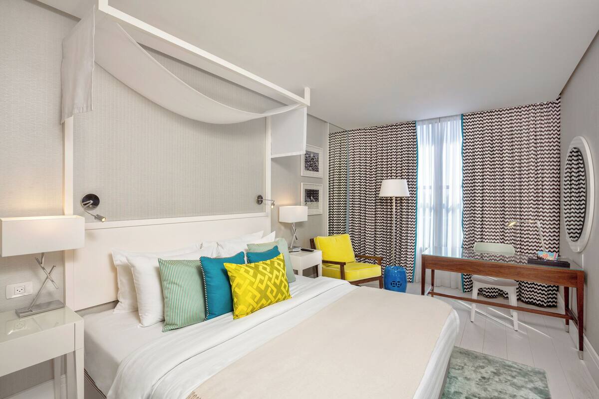 Quarto de hotel com cama, mesas de cabeceira, escrivaninha, cadeira e espelho.