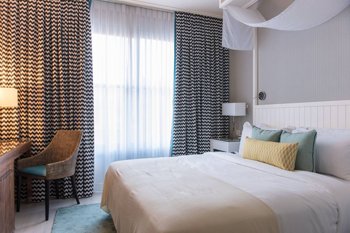 Quarto de hotel com cama, cadeira, mesa de cabeceira e cortinas.