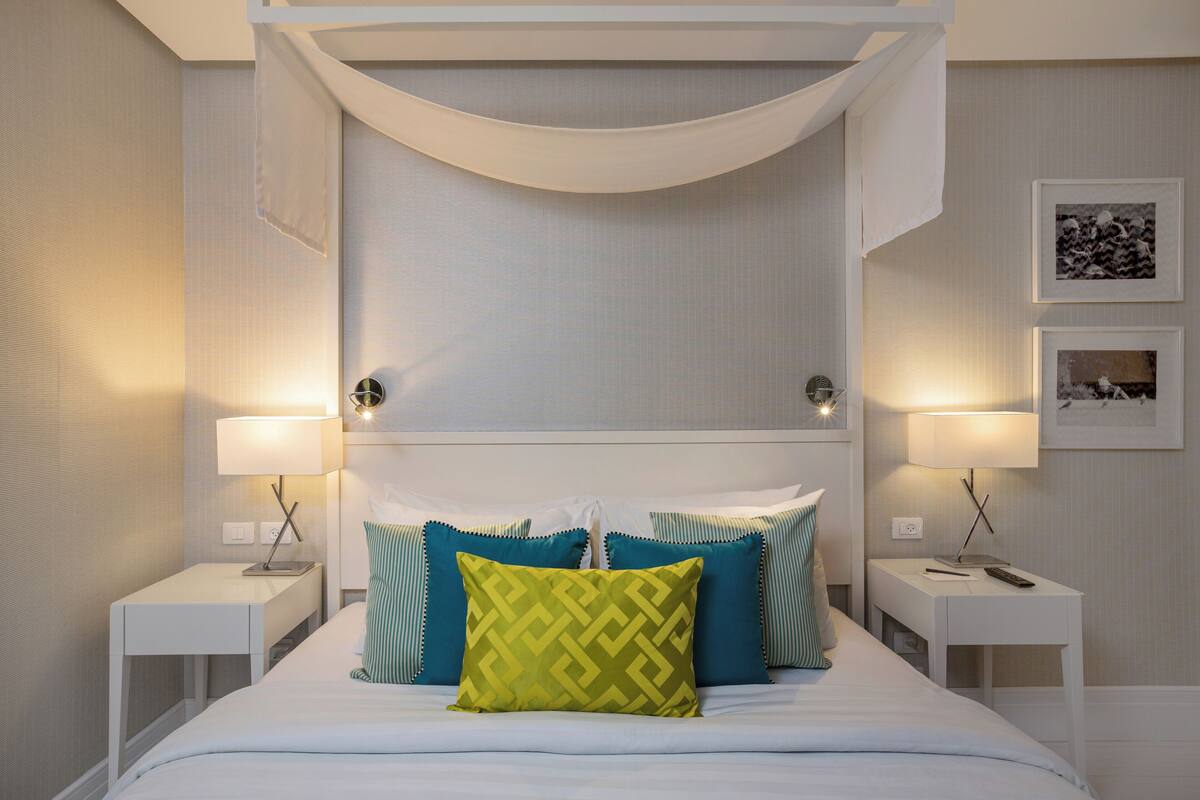 Quarto de hotel bem arrumado com uma cama, duas mesas de cabeceira e duas luminárias fixadas na parede.