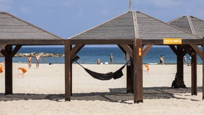 Uma praia com cabanas de madeira, uma rede e cadeiras laranja.