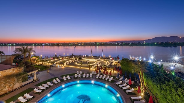 Área da piscina com espreguiçadeiras e guarda-sóis, rodeada por um jardim bem cuidado e vista para a água.