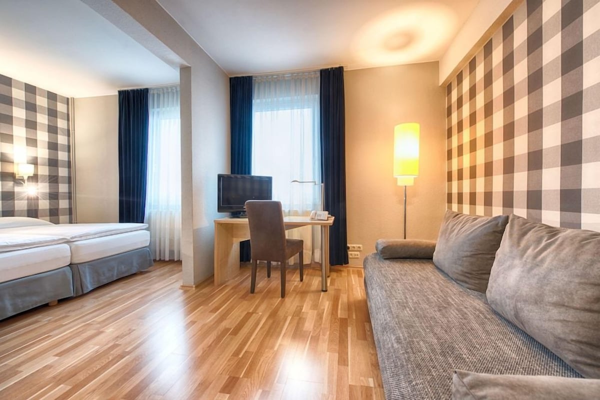 Quarto de hotel com cama, escrivaninha com computador, sofá e abajur.