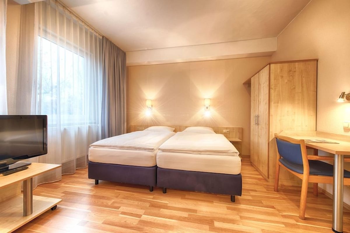 Quarto de hotel com duas camas, televisão, escrivaninha e guarda-roupa.