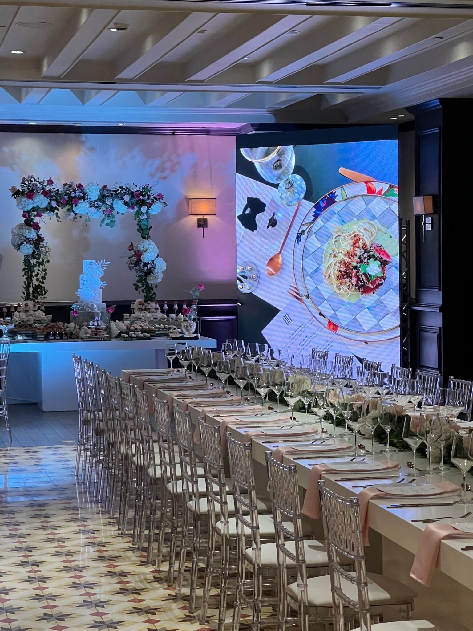 Salão de banquetes com mesas longas preparadas para um evento formal, apresentando um arco floral e uma grande tela digital exibindo um prato.