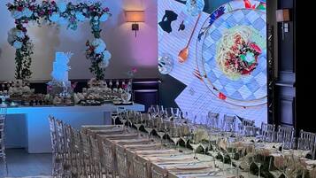 Salão de banquetes com mesas longas preparadas para um evento formal, apresentando um arco floral e uma grande tela digital exibindo um prato.