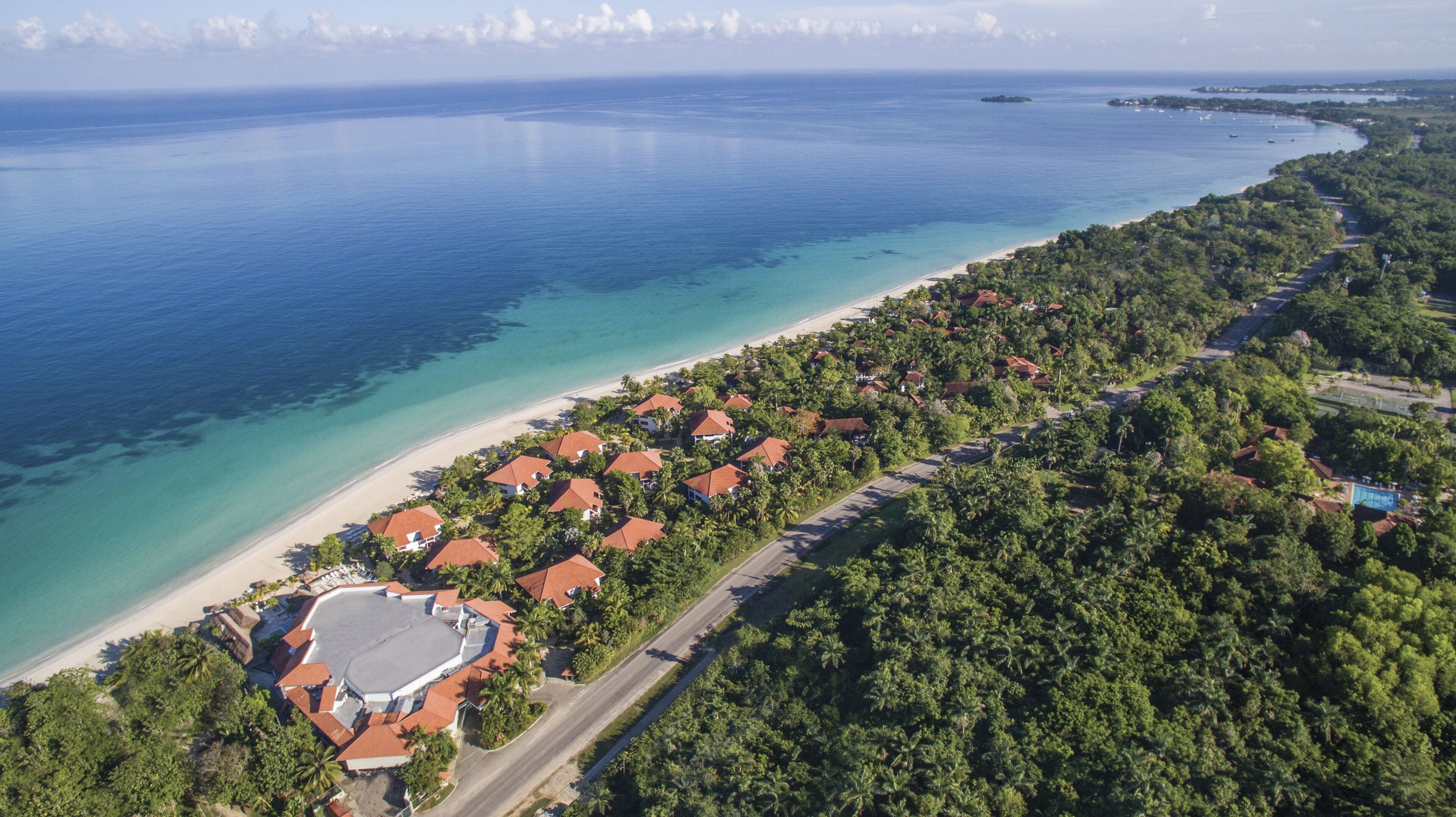 Vista aérea de uma estância costeira com praia, vegetação densa e mar azul cristalino.
