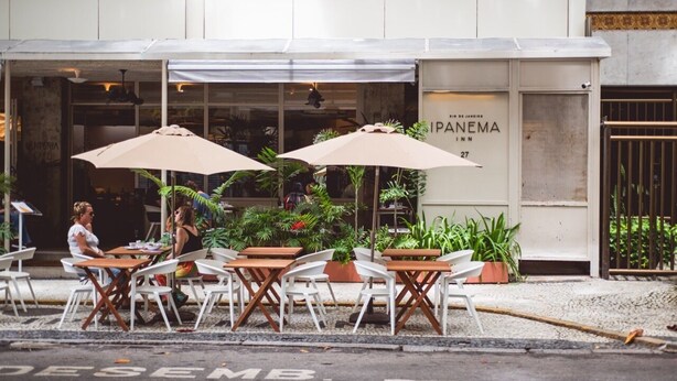 Uma área de estar ao ar livre com mesas, cadeiras, guarda-sóis e plantas em vasos.