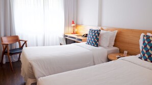 Quarto de hotel com duas camas, uma escrivaninha, uma cadeira e uma mesa de cabeceira com abajur.