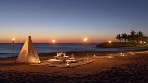 Uma praia com uma tenda, uma área de refeições e um afloramento rochoso com palmeiras.
