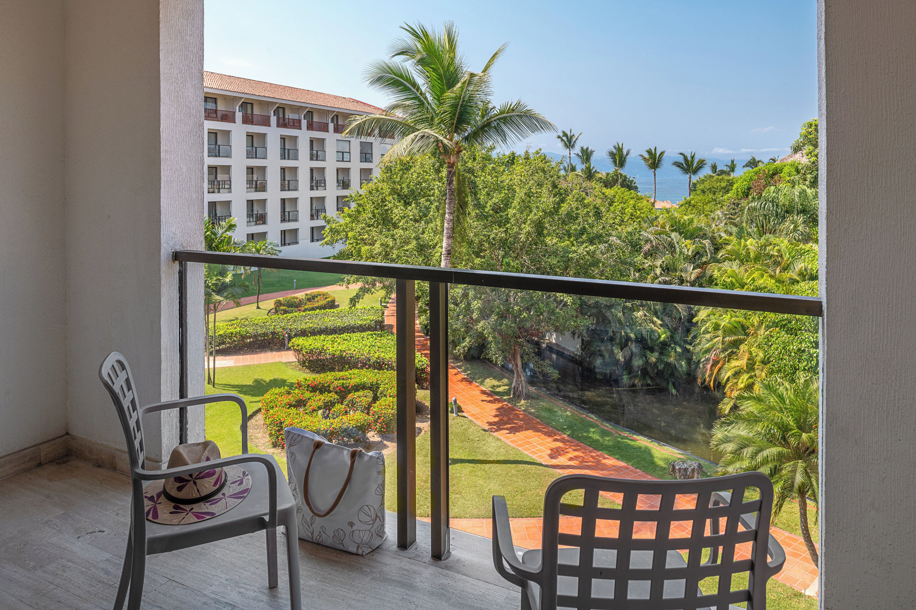 Uma varanda com duas cadeiras, uma mesa pequena e vista para um jardim ajardinado e um edifício ao fundo.
