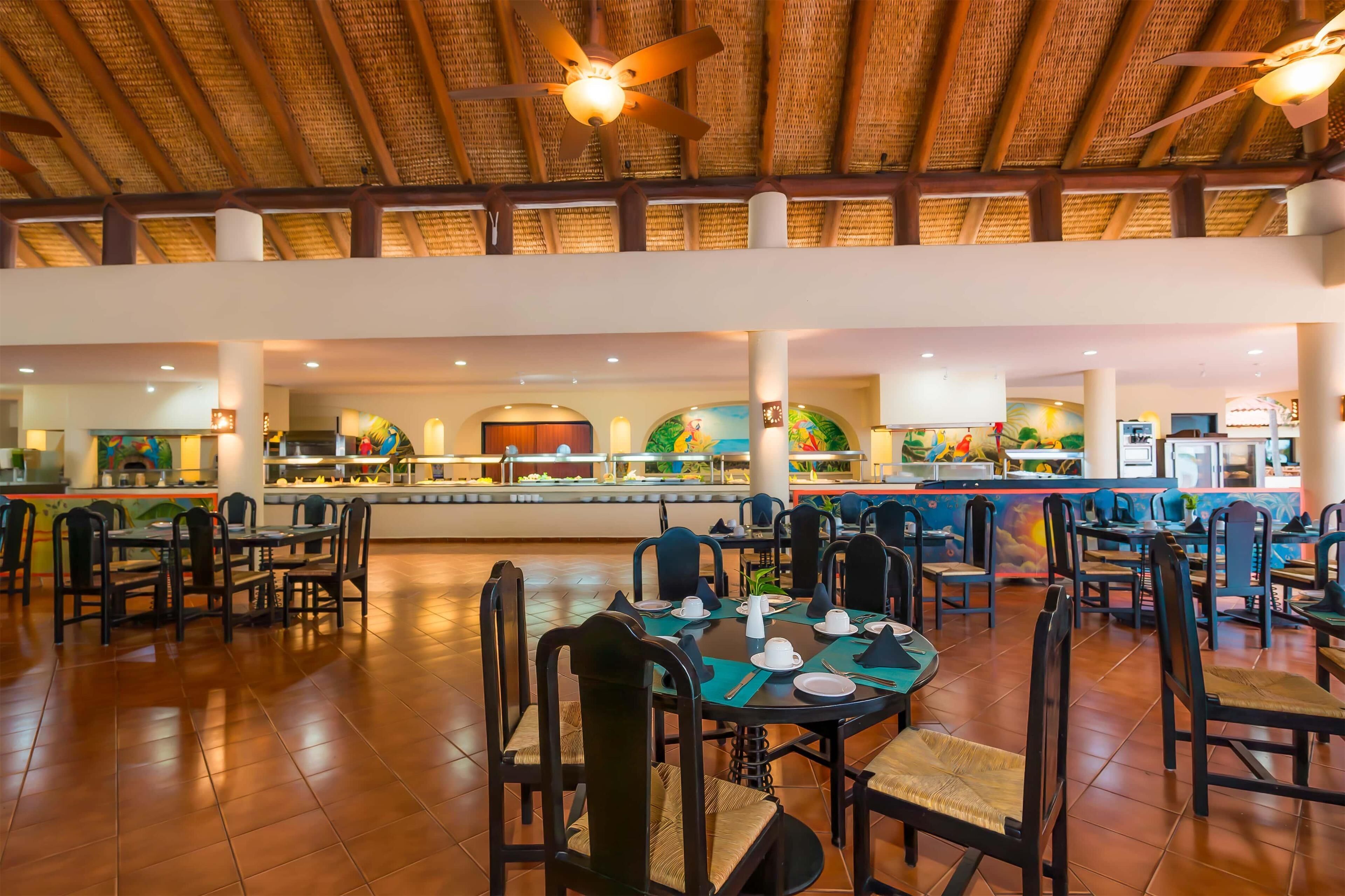 Uma sala de jantar ampla com pé direito alto e teto de palha, com várias mesas e cadeiras, e um balcão de buffet ao fundo.