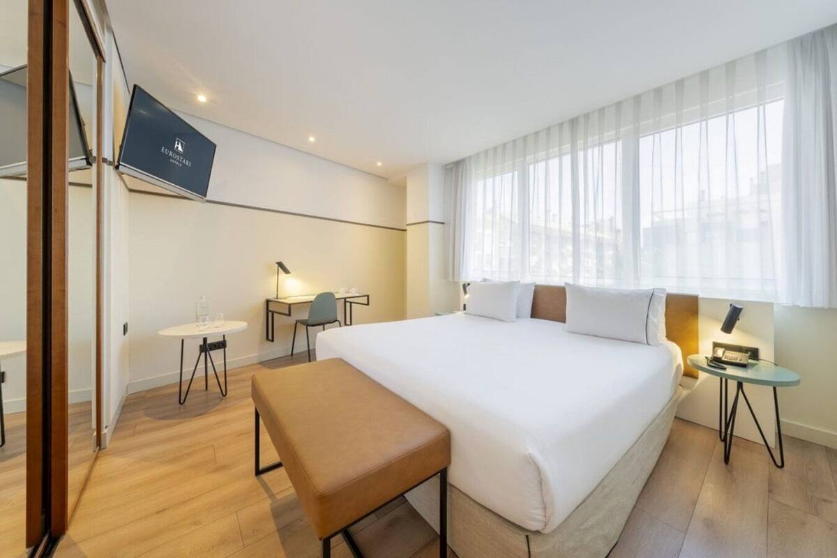 Quarto de hotel moderno com uma cama grande, uma escrivaninha, uma cadeira, uma mesinha e uma televisão fixada na parede.