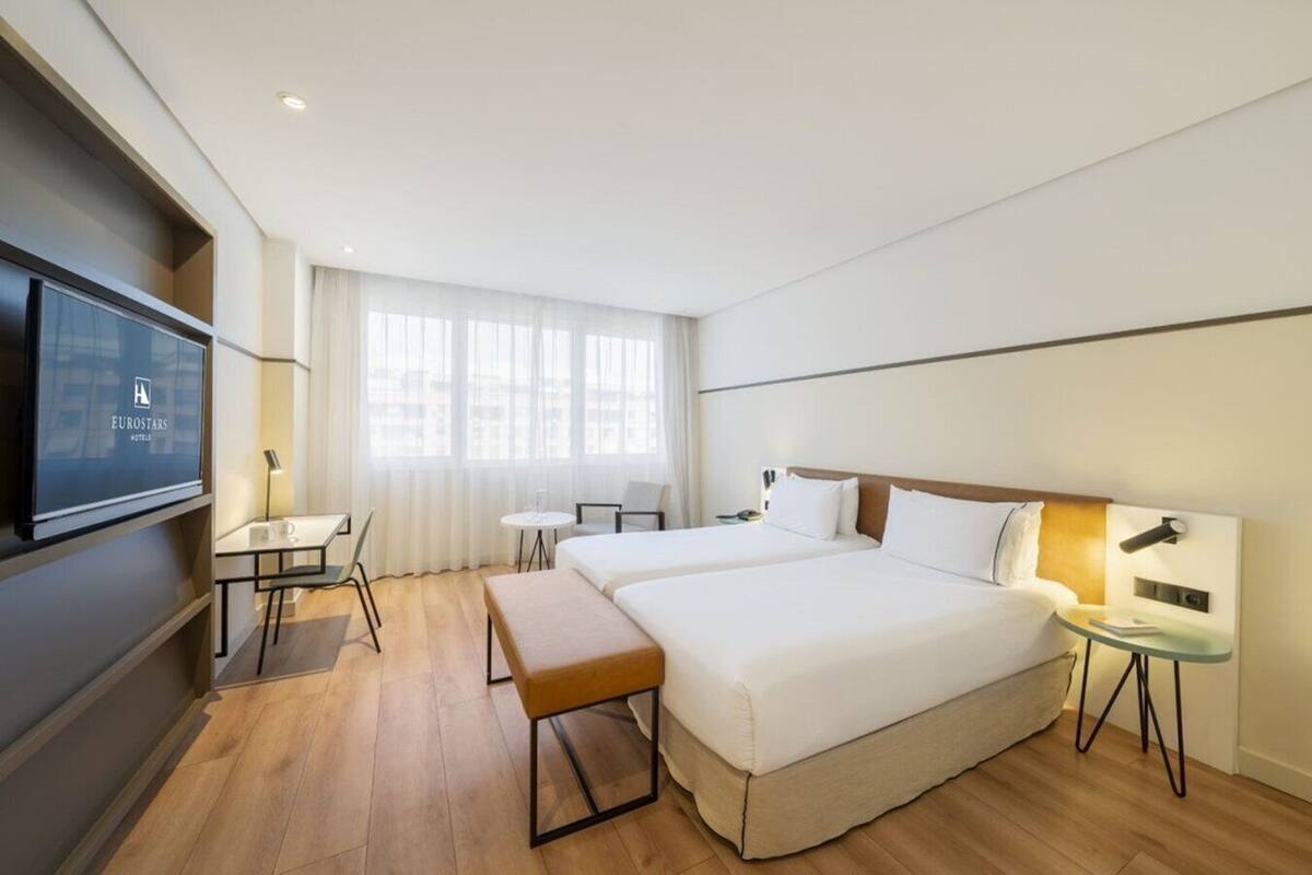 Quarto de hotel com cama, escrivaninha, cadeira, televisão e janela com cortinas.