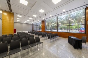 Uma sala de conferências com fileiras de cadeiras, um pódio e amplas janelas com vista para palmeiras e edifícios.
