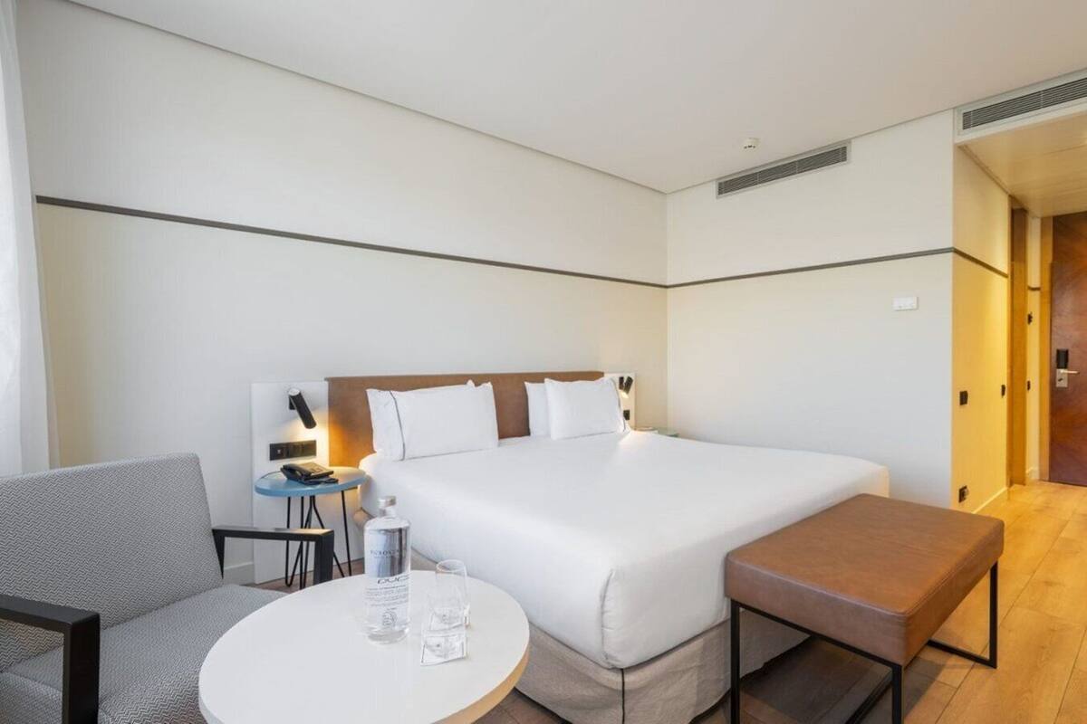 Quarto de hotel com uma cama grande, uma mesinha de cabeceira com abajur, uma cadeira e uma mesa redonda pequena com copos e uma garrafa.