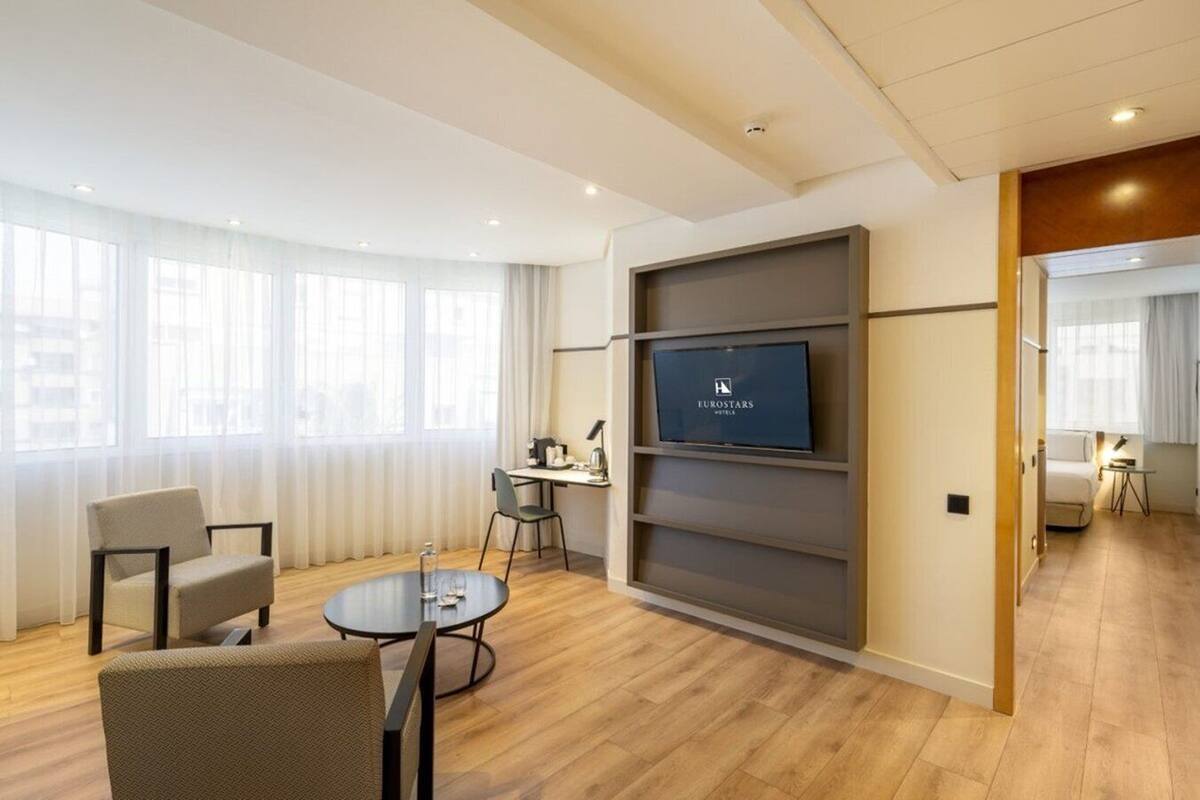 Quarto de hotel moderno com uma TV grande na parede, uma mesa de jantar redonda, duas cadeiras, uma escrivaninha com cadeira e uma janela ampla com cortinas translúcidas.