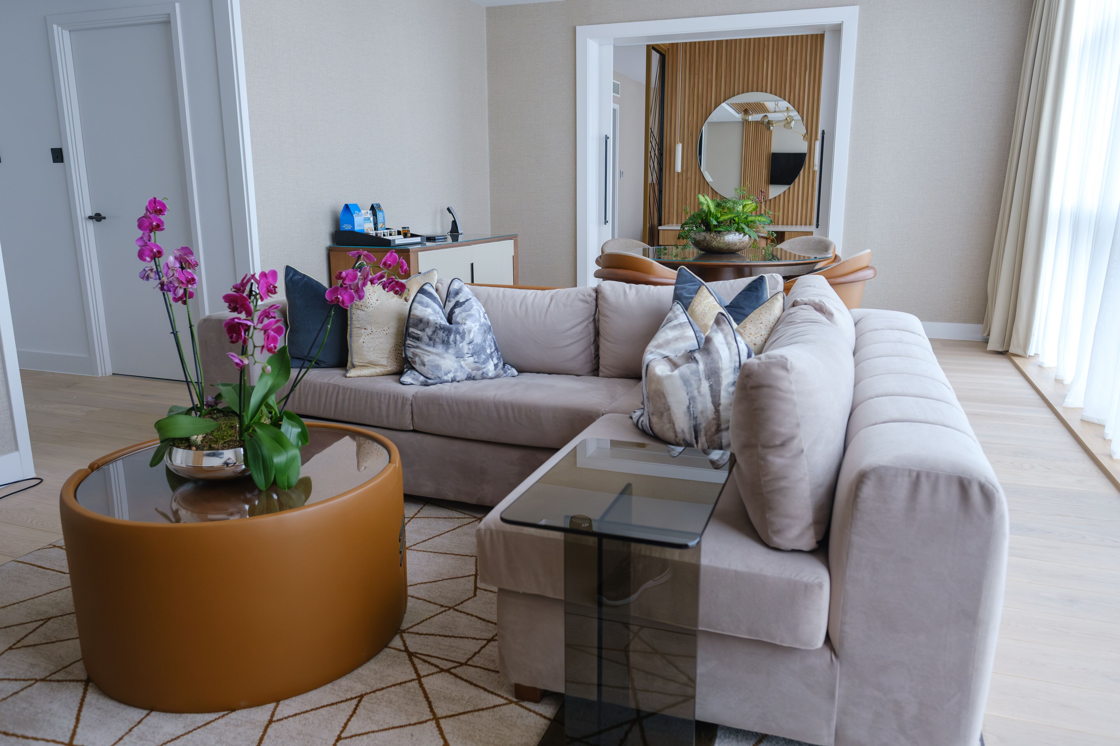 Uma sala de estar moderna com um sofá bege, uma mesa de centro de vidro e um cachepot laranja redondo com orquídeas. Há um aparador de madeira com um espelho e uma planta em vaso.