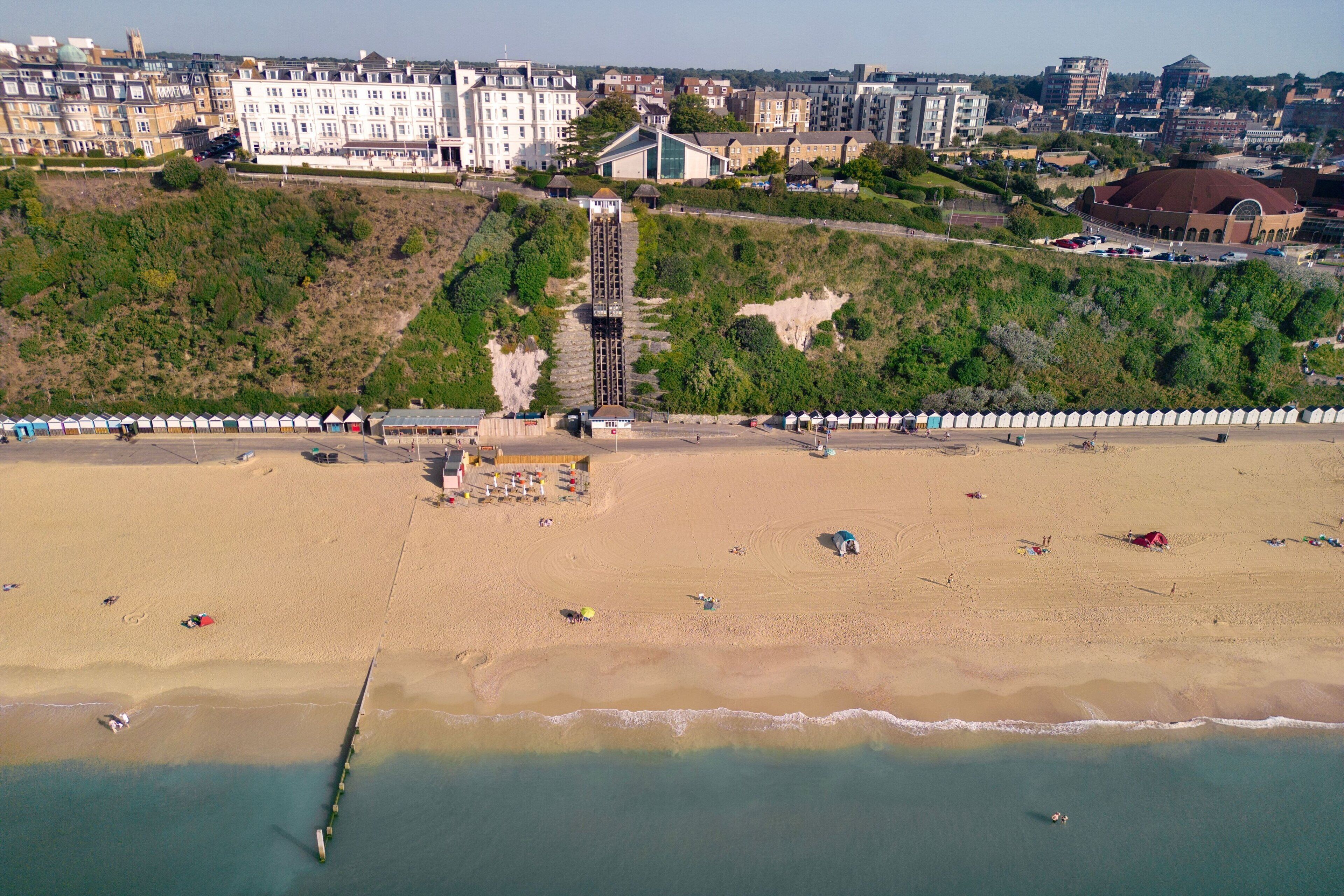 Vista aérea de uma praia com um cais longo que se estende para a água, uma fileira de cabanas de praia e um edifício grande com telhado em cúpula ao fundo.