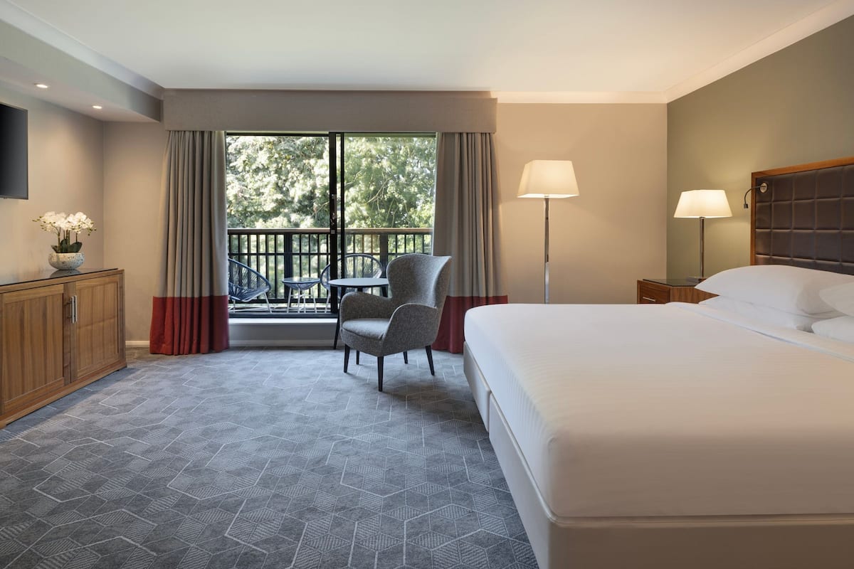 Quarto de hotel com uma cama grande, uma escrivaninha, uma cadeira, uma televisão e uma varanda com cortinas.