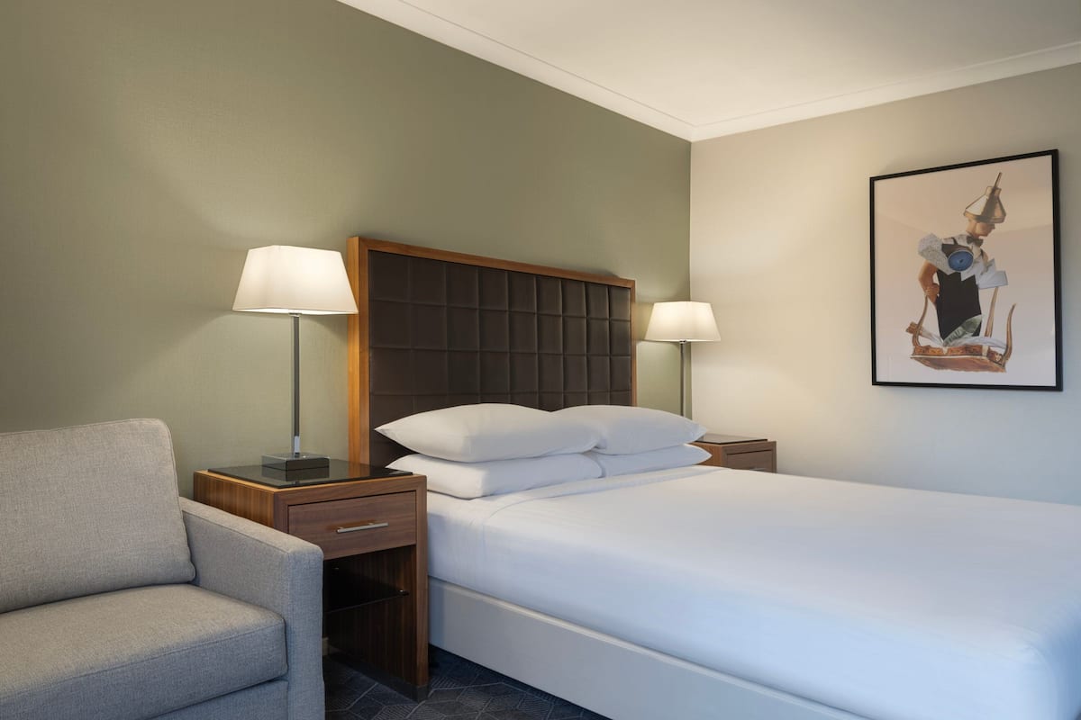 Quarto de hotel com uma cama, duas luminárias de cabeceira, uma poltrona, mesa de cabeceira e uma obra de arte emoldurada na parede.