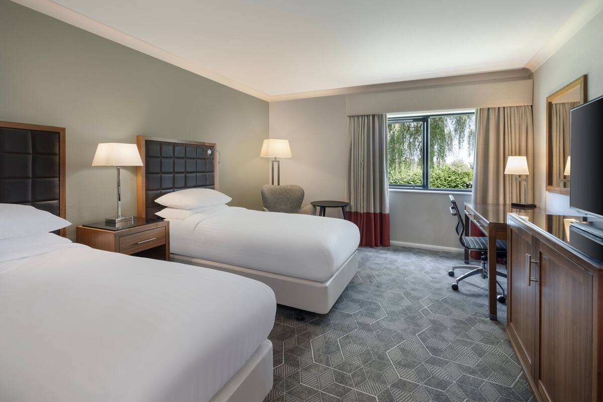 Quarto de hotel com duas camas, uma mesa, uma cadeira, uma televisão e uma janela com cortinas.