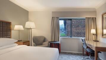 Quarto de hotel com uma cama, uma escrivaninha, uma cadeira, duas luminárias e uma janela com cortinas.