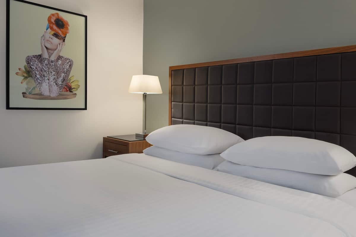 Cama bem arrumada com lençóis brancos, cabeceira escura, mesa de cabeceira com abajur e uma obra de arte emoldurada na parede.
