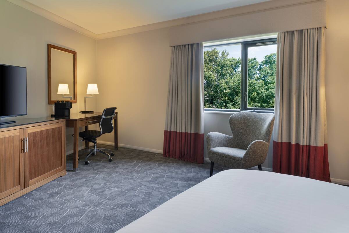Quarto de hotel com cama, escrivaninha com computador e cadeira, televisão e janela com cortinas.